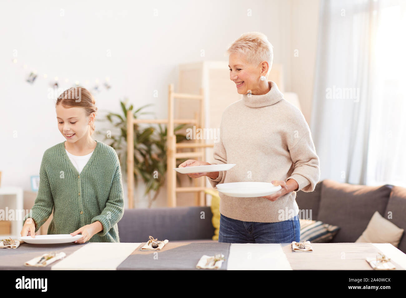 Smiling mature mother putting plates on the table with her daughter ...