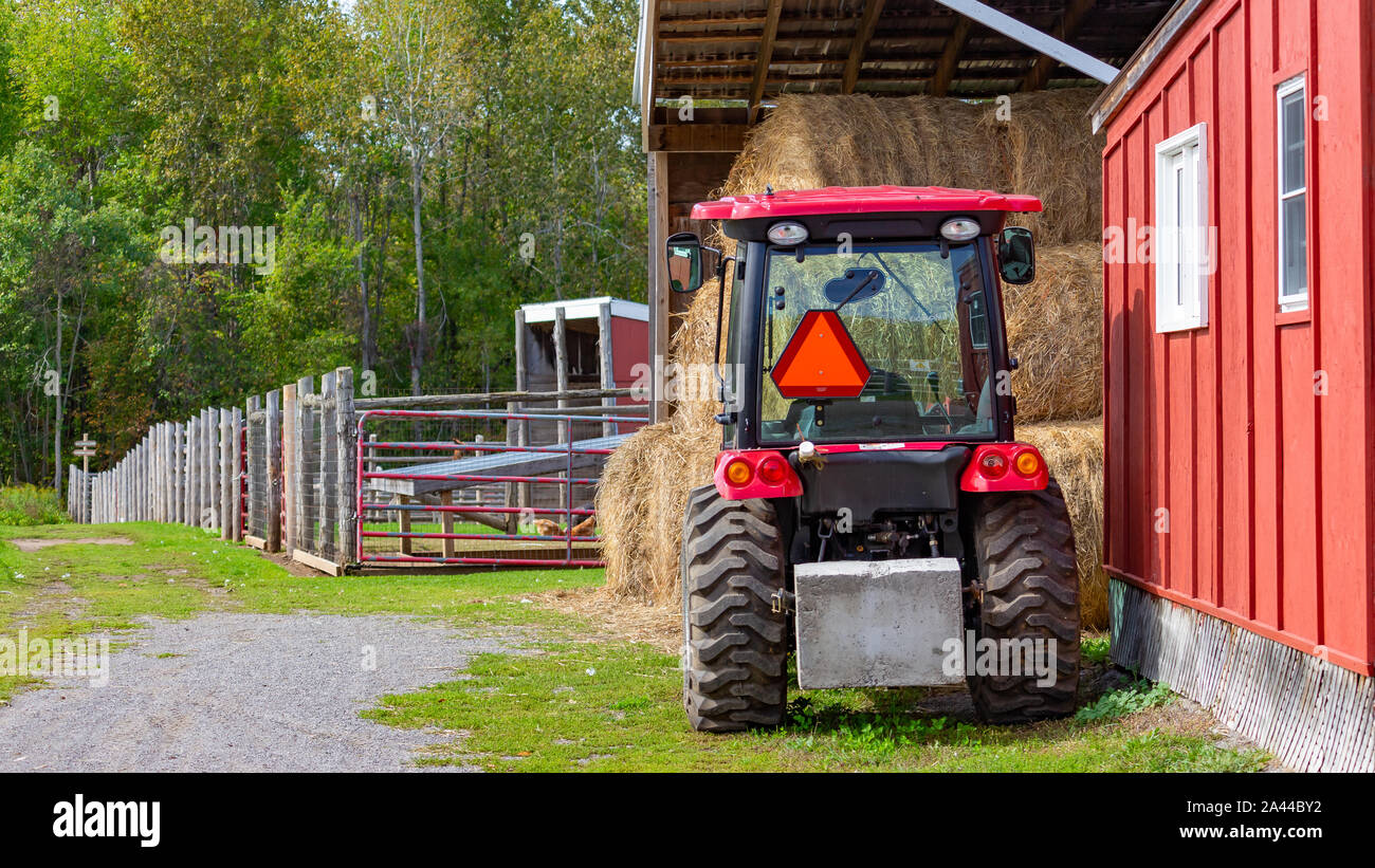 A small, red tractor is parked beside a barn and bales of hay on a farm ...