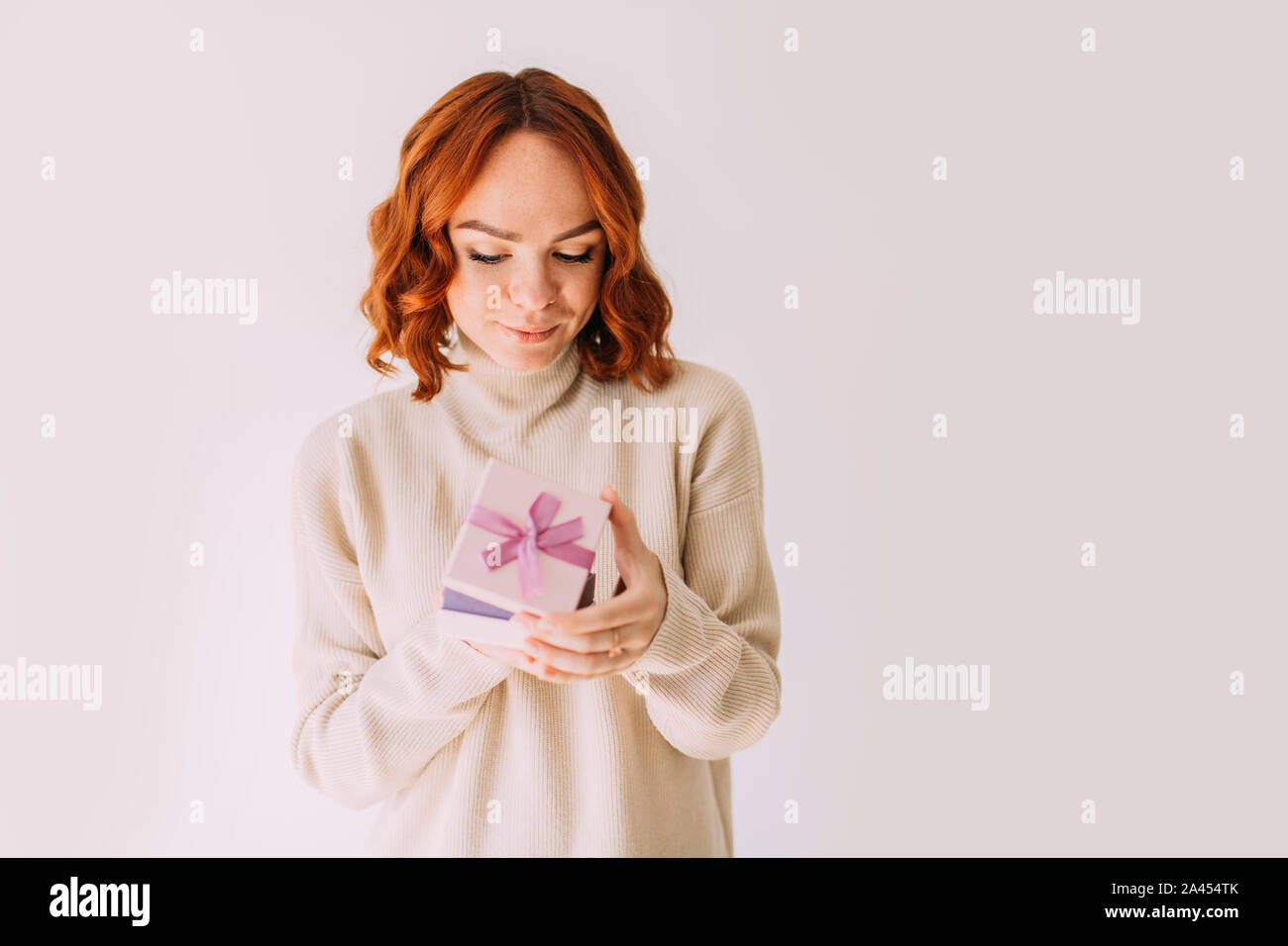 Red-haired festive girl smiles, holding a pastel coloured gift box with ...