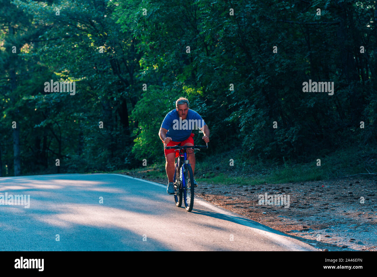 Older cyclist in his mid-40s riding a bike on a road through the woods ...