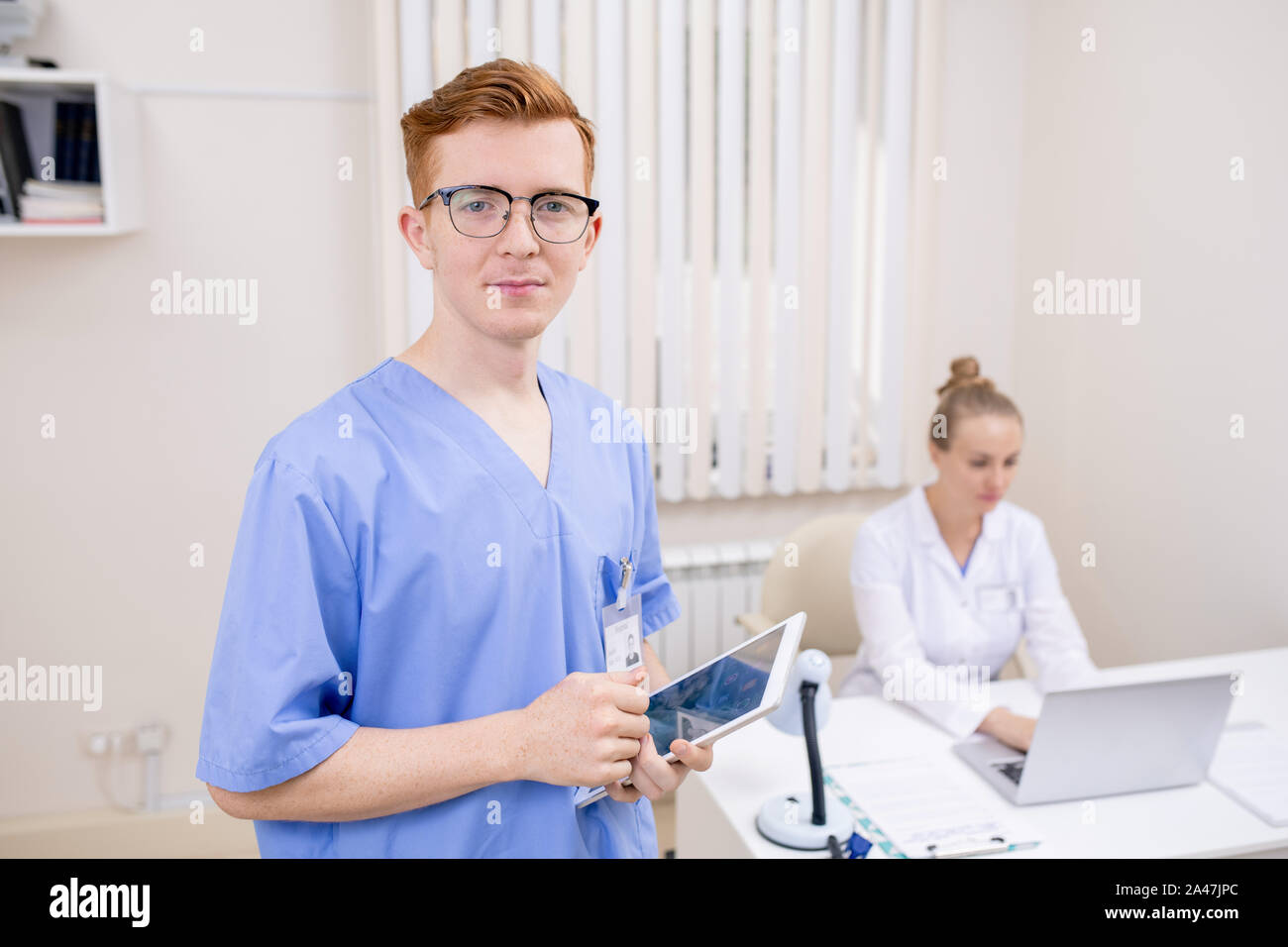 Young mobile intern in eyeglasses and blue medical uniform using ...