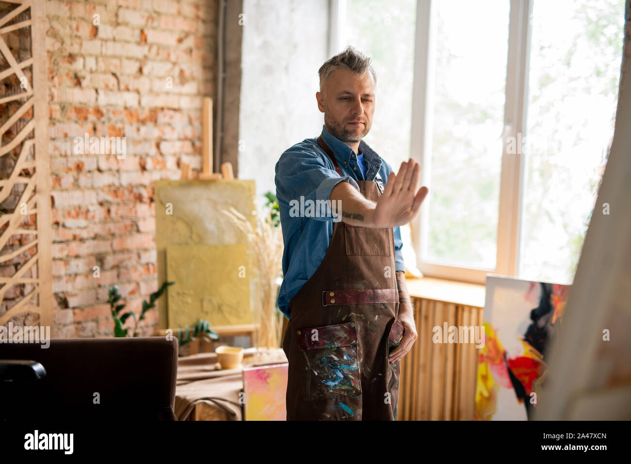 Creative painter in workwear covering part of painting with hand Stock ...