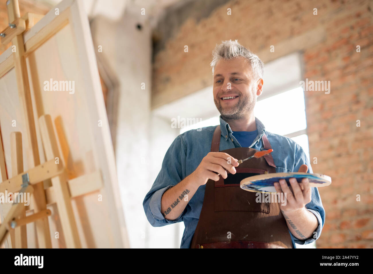 Happy and successful painter with palette looking at painting on easel ...