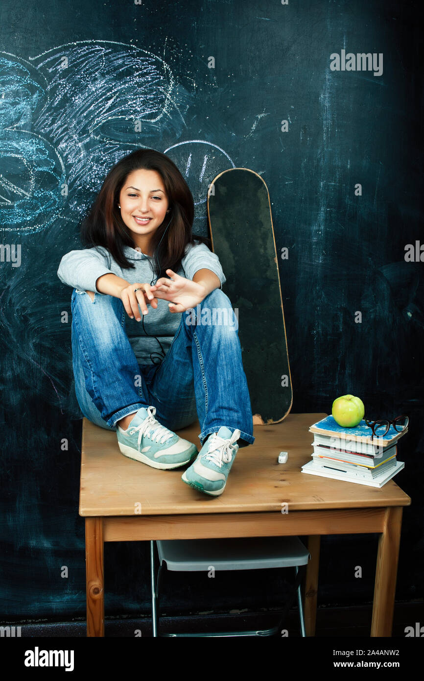 young cute teenage girl in classroom at blackboard seating on table ...