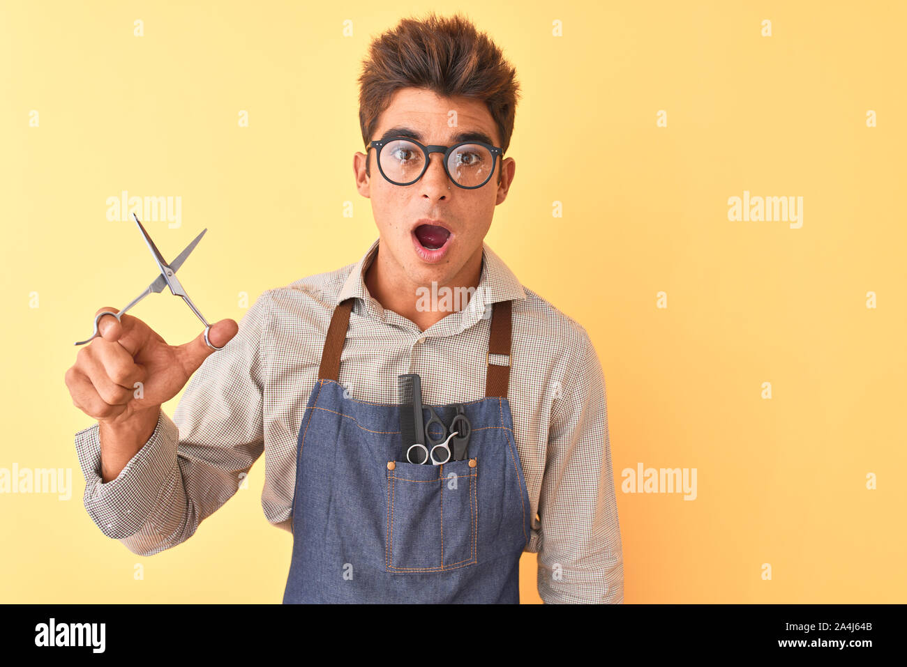 Handsome hairdresser man wearing apron holding scissors over isolated yellow background scared ...