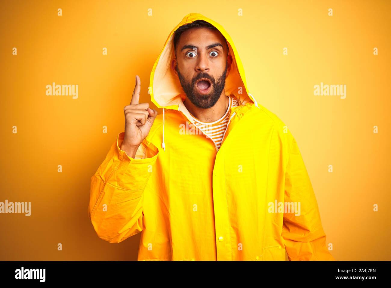 Young indian man wearing raincoat with hood standing over isolated yellow background pointing ...