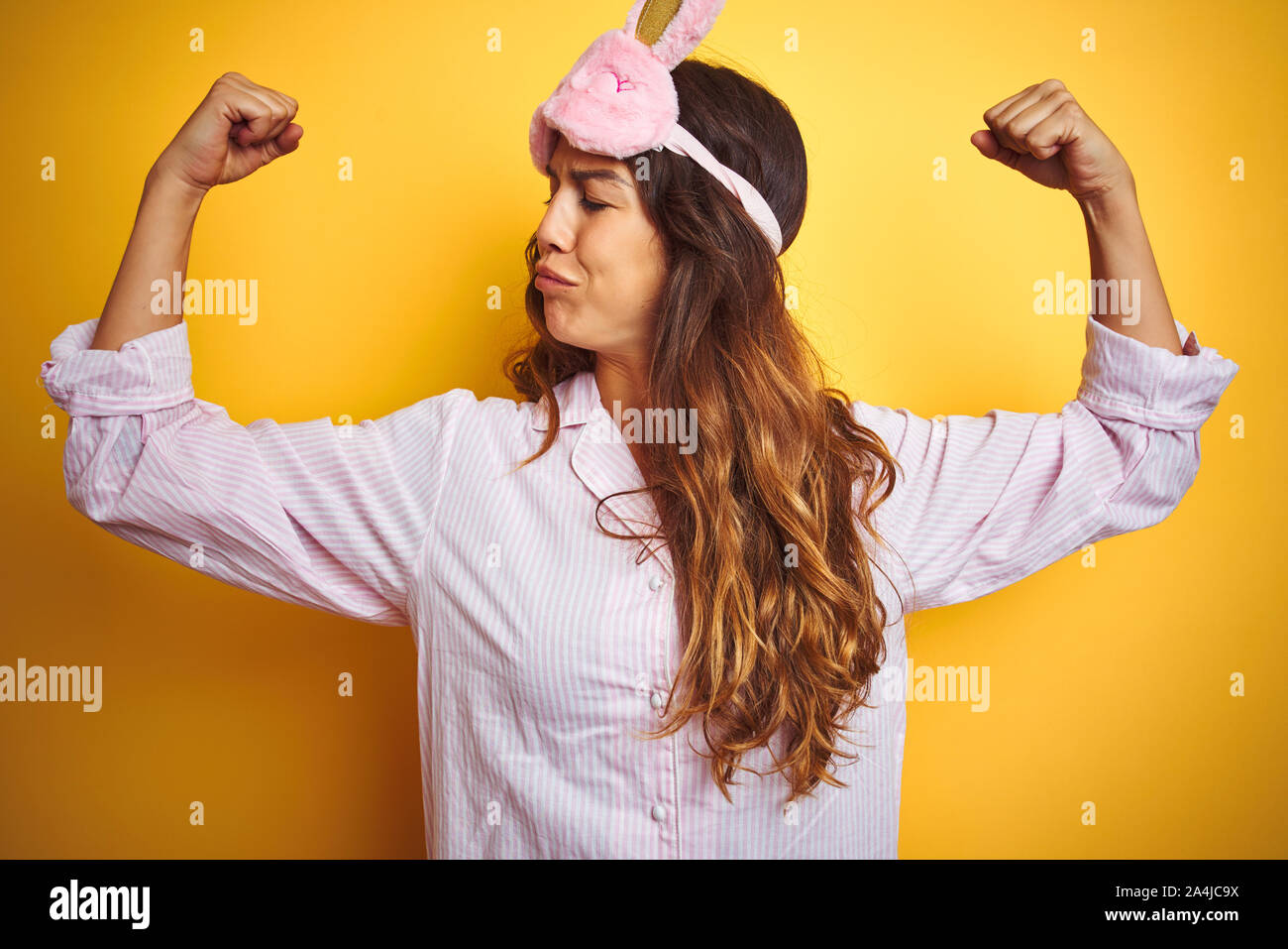 Young woman wearing pajama and sleep mask standing over yellow isolated ...