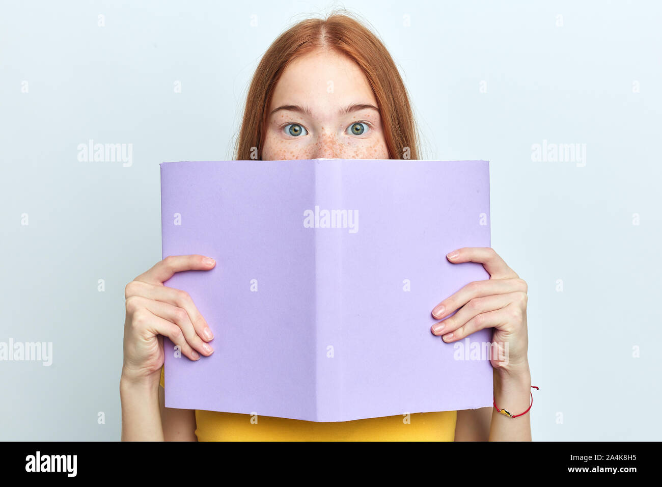 red-headed scared woman closing her face with a copy book, looking at ...