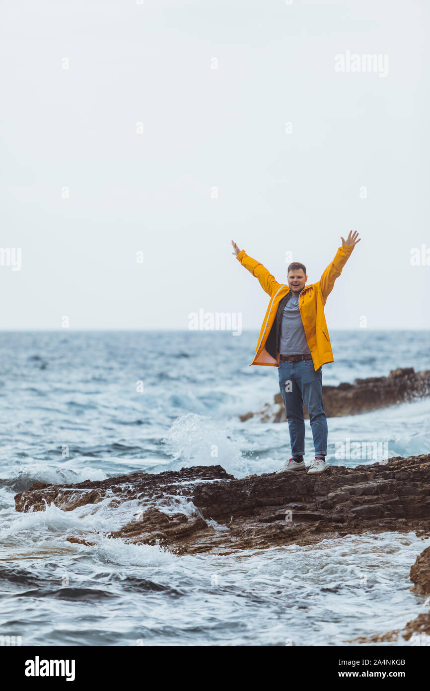 young man rise up hands at rocky sea coast in yellow raincoat in storm ...