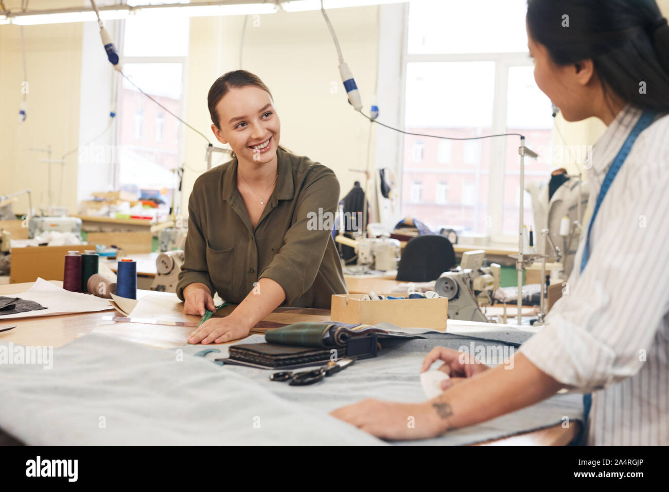 Happy young designer smiling to her colleague while they sewing new ...