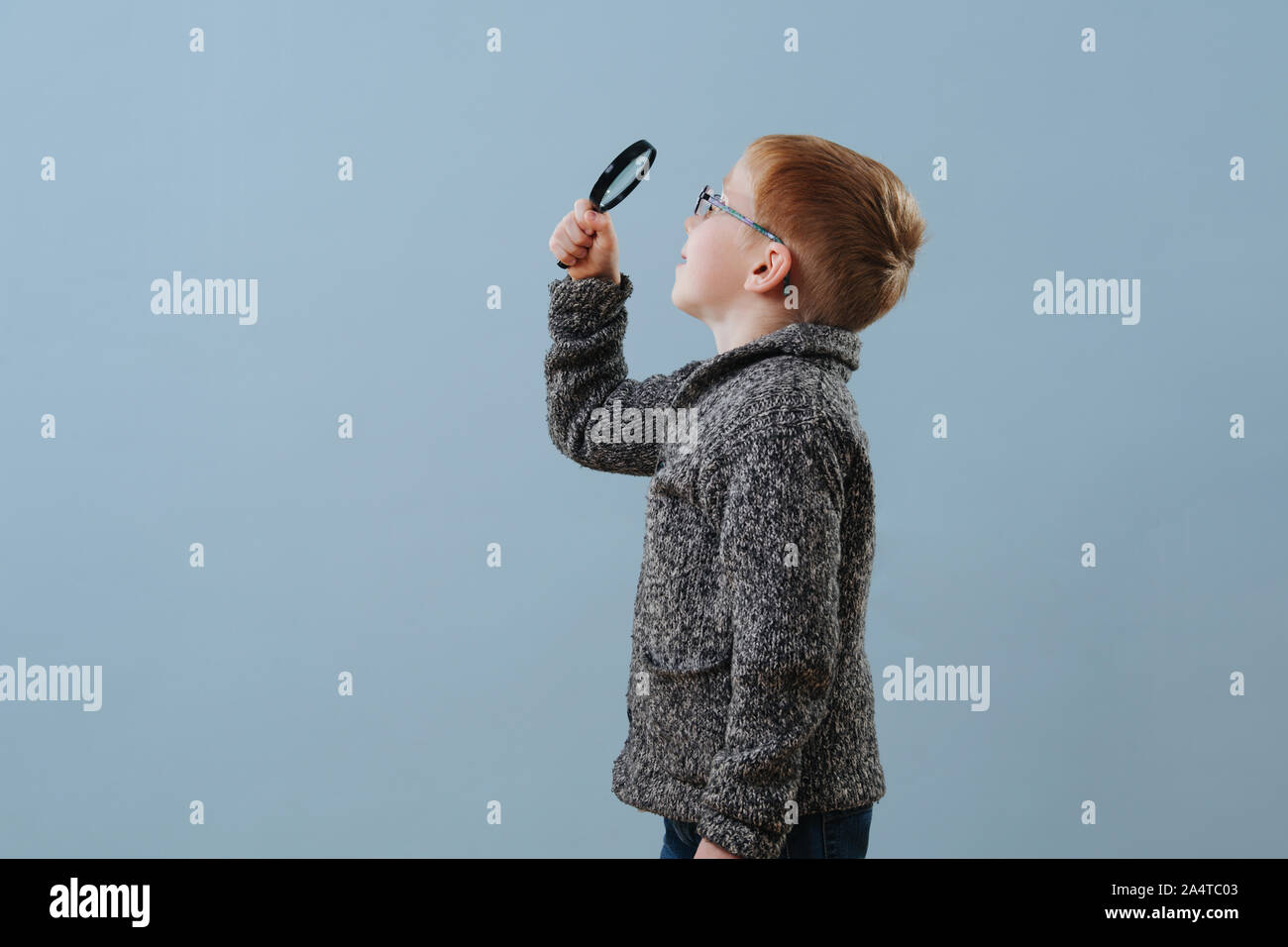 Curious ginger boy in glasses looking up through magnifying glass over ...