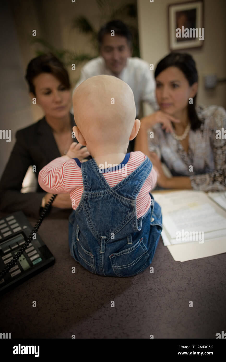 Young baby sitting on a desk in front of three businesspeople inside an ...
