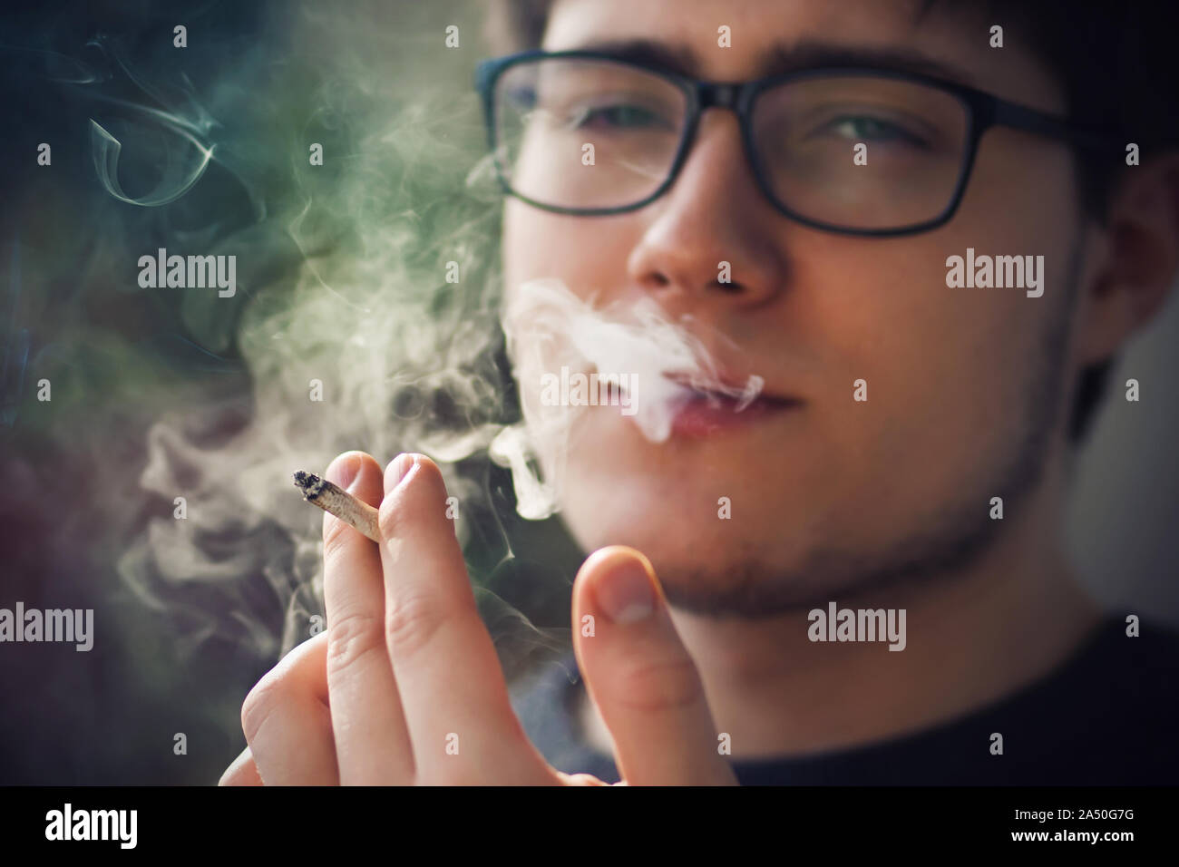 A young handsome guy in glasses smokes a cigarette and exhales smoke ...