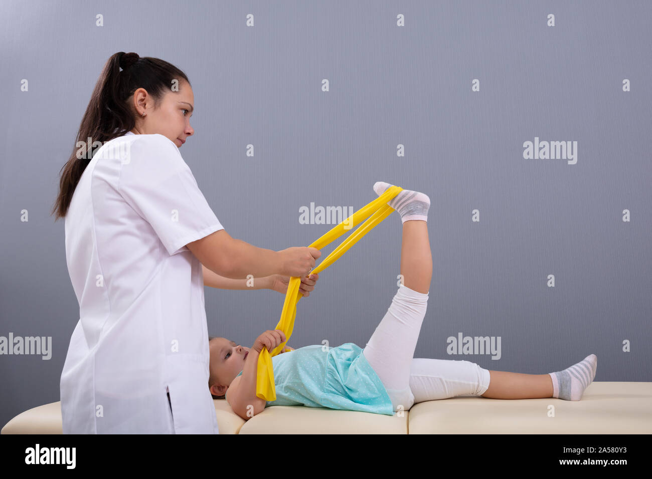 Girl Stretching The Leg With Yellow Elastic Band Instructed By A Female ...