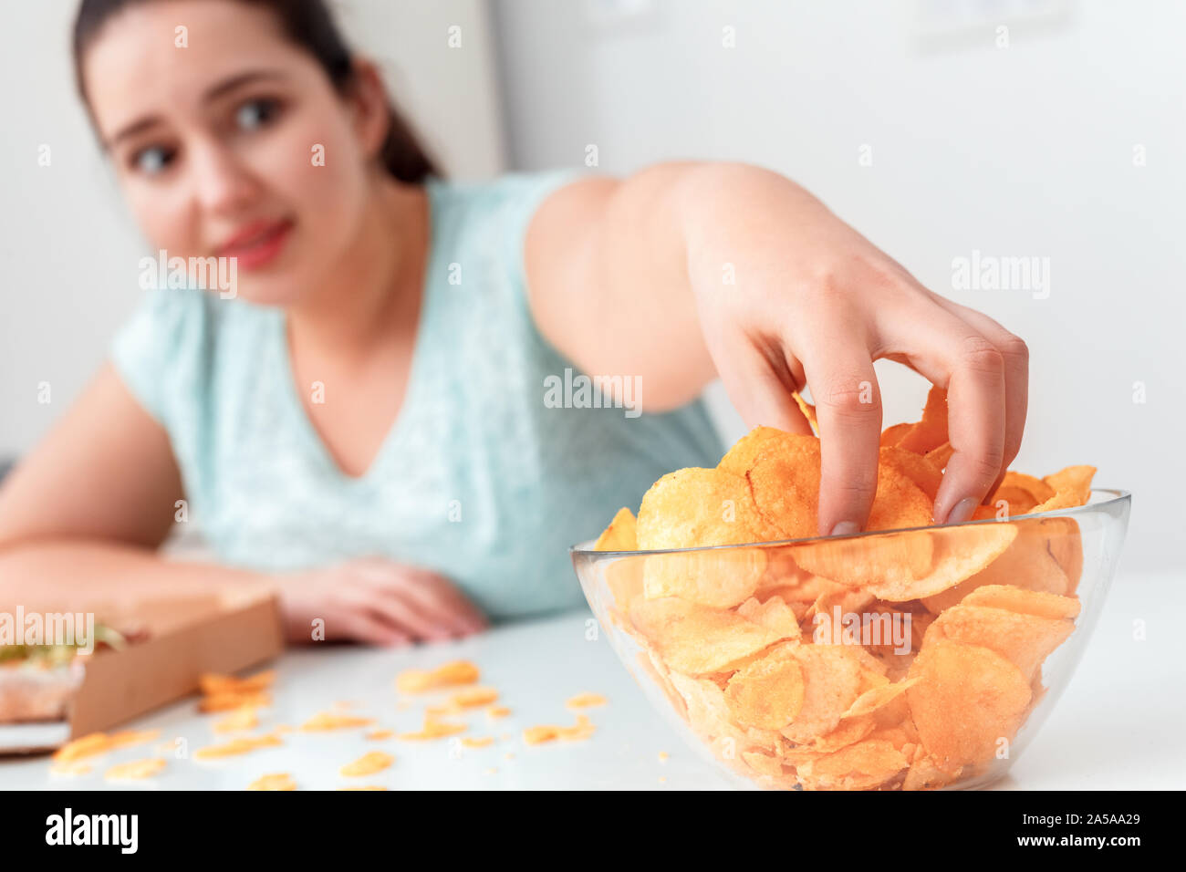 Breaking Diet. Chubby girl sitting at kitchen table eating chips ...