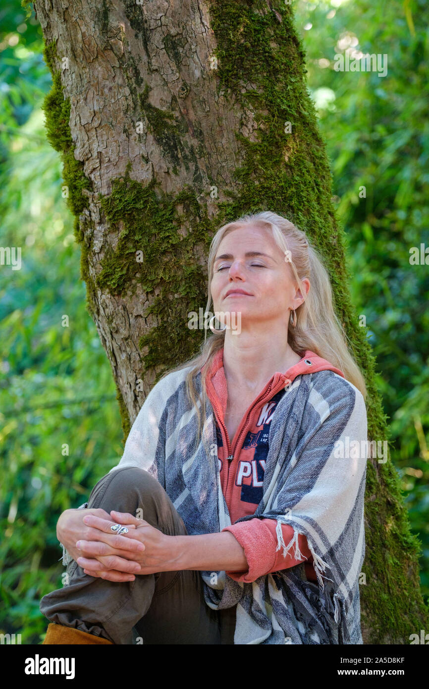 Blond woman sitting next to a tree with eyes closed during a forest ...