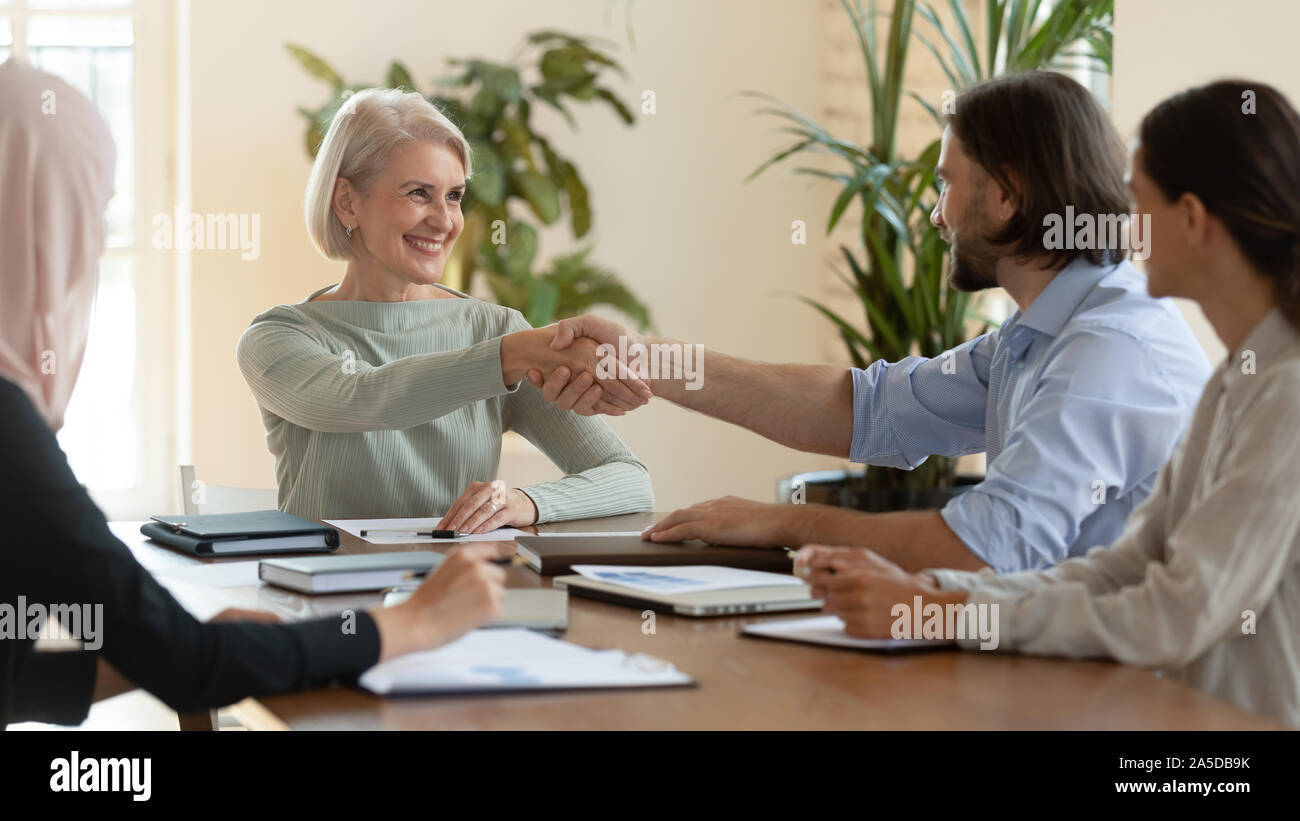 Smiling mature female leader handshake male partner at team meeting ...