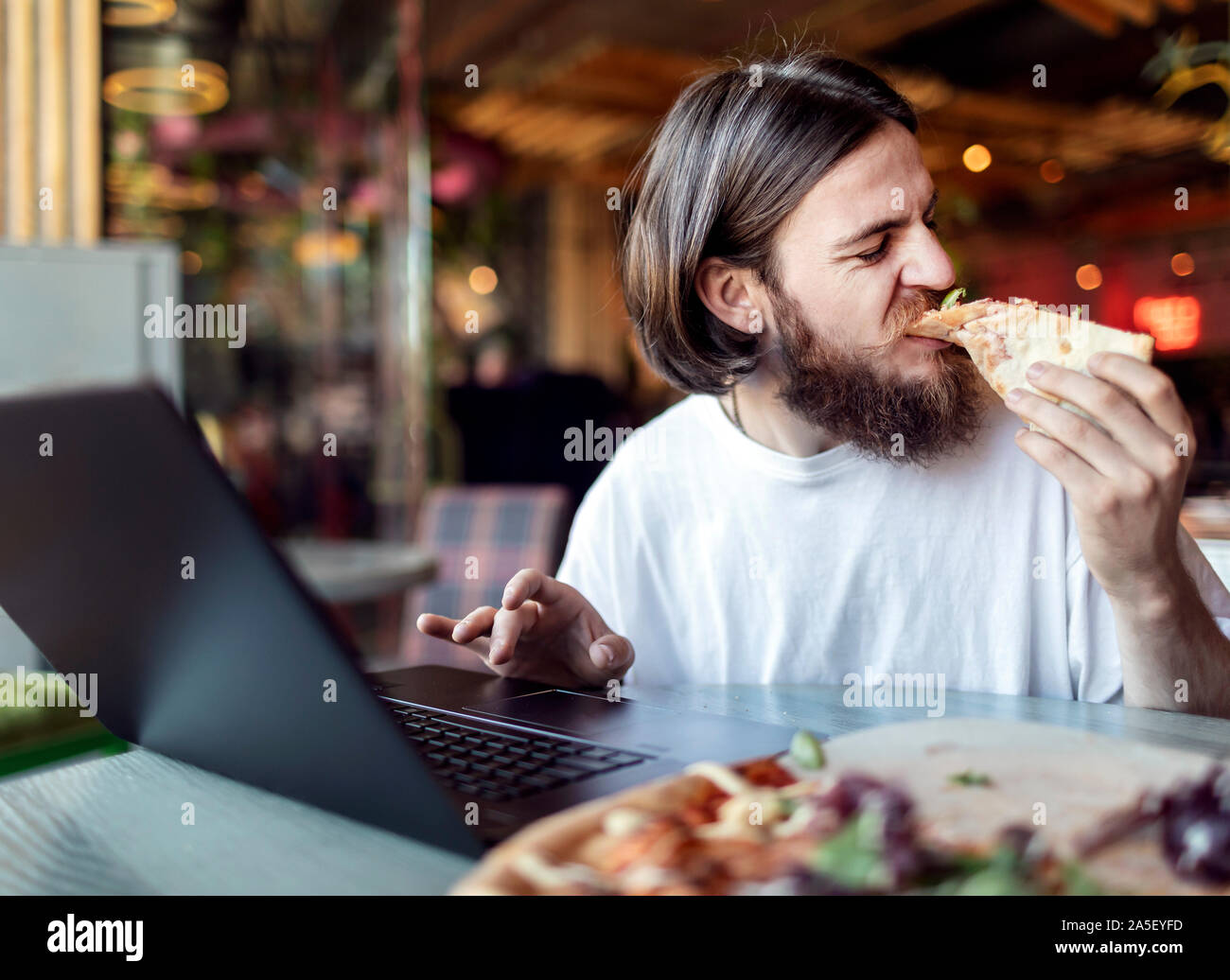 Young freelancer male snacking while working by laptop in a summer cafe ...