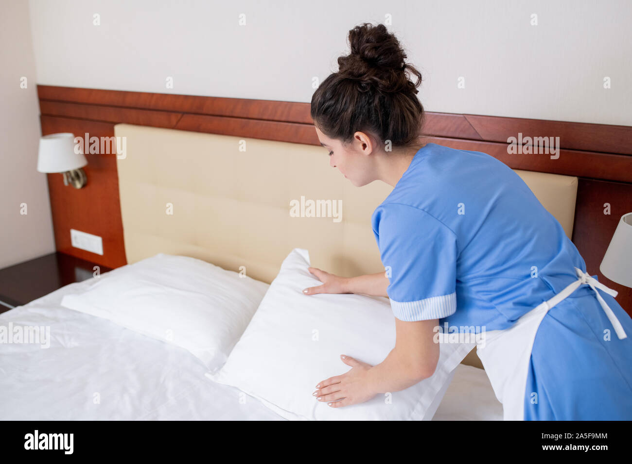 Young brunette female in blue uniform and white apron bending over bed ...
