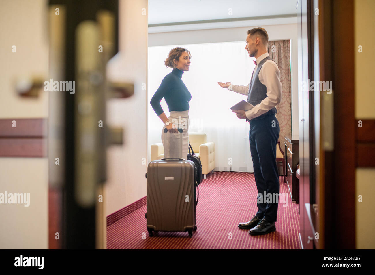 Pretty young elegant woman with luggage standing in hotel room and ...