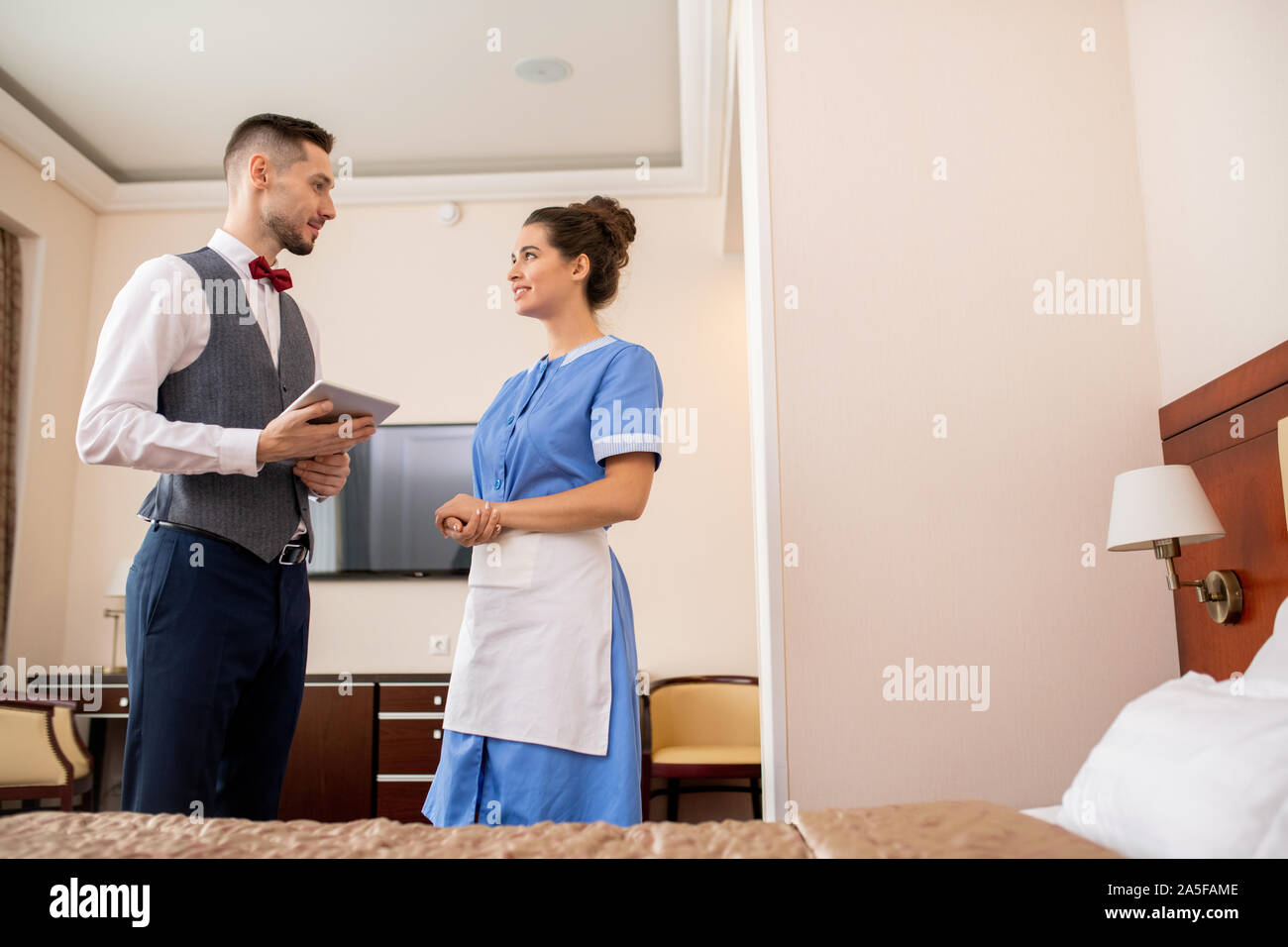 Young elegant porter with touchpad looking at pretty chamber maid in ...