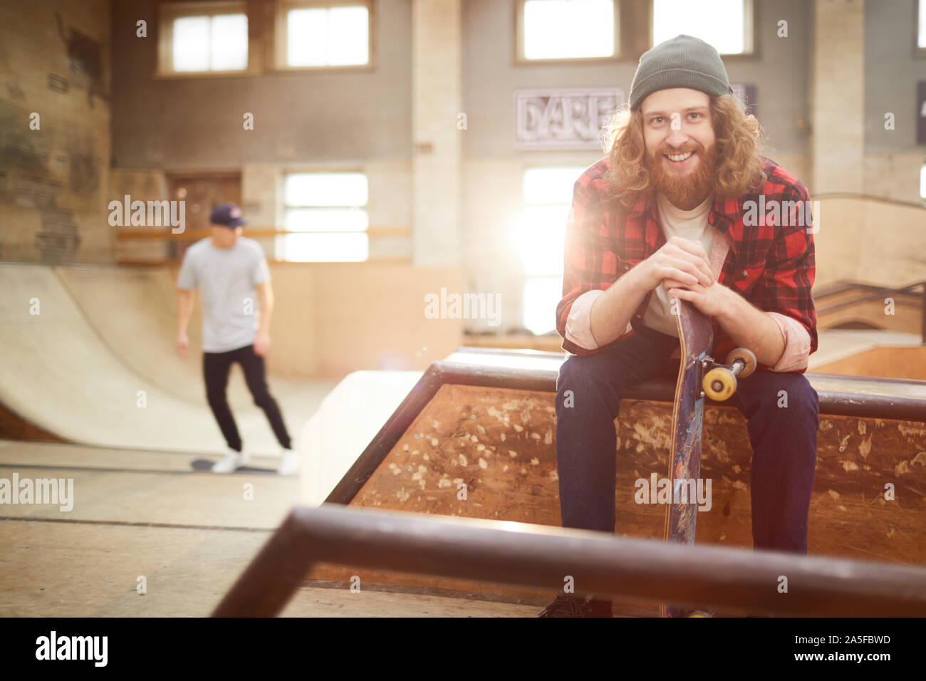 Portrait of cheerful bearded skater looking at camera while siting on ...