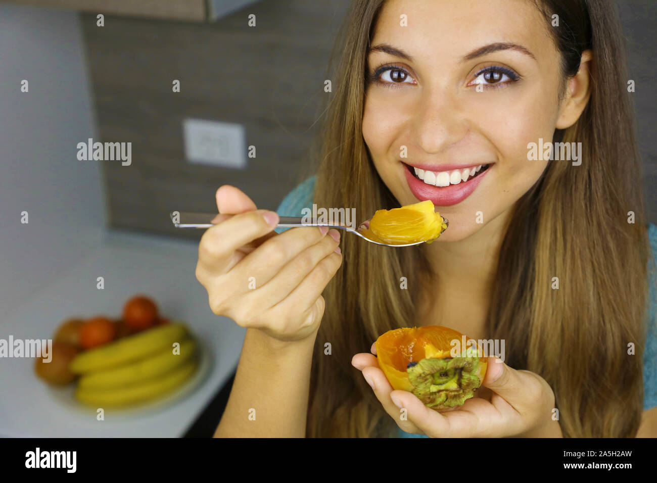 Beautiful young woman smiling and eating persimmon kaki fruit at home ...