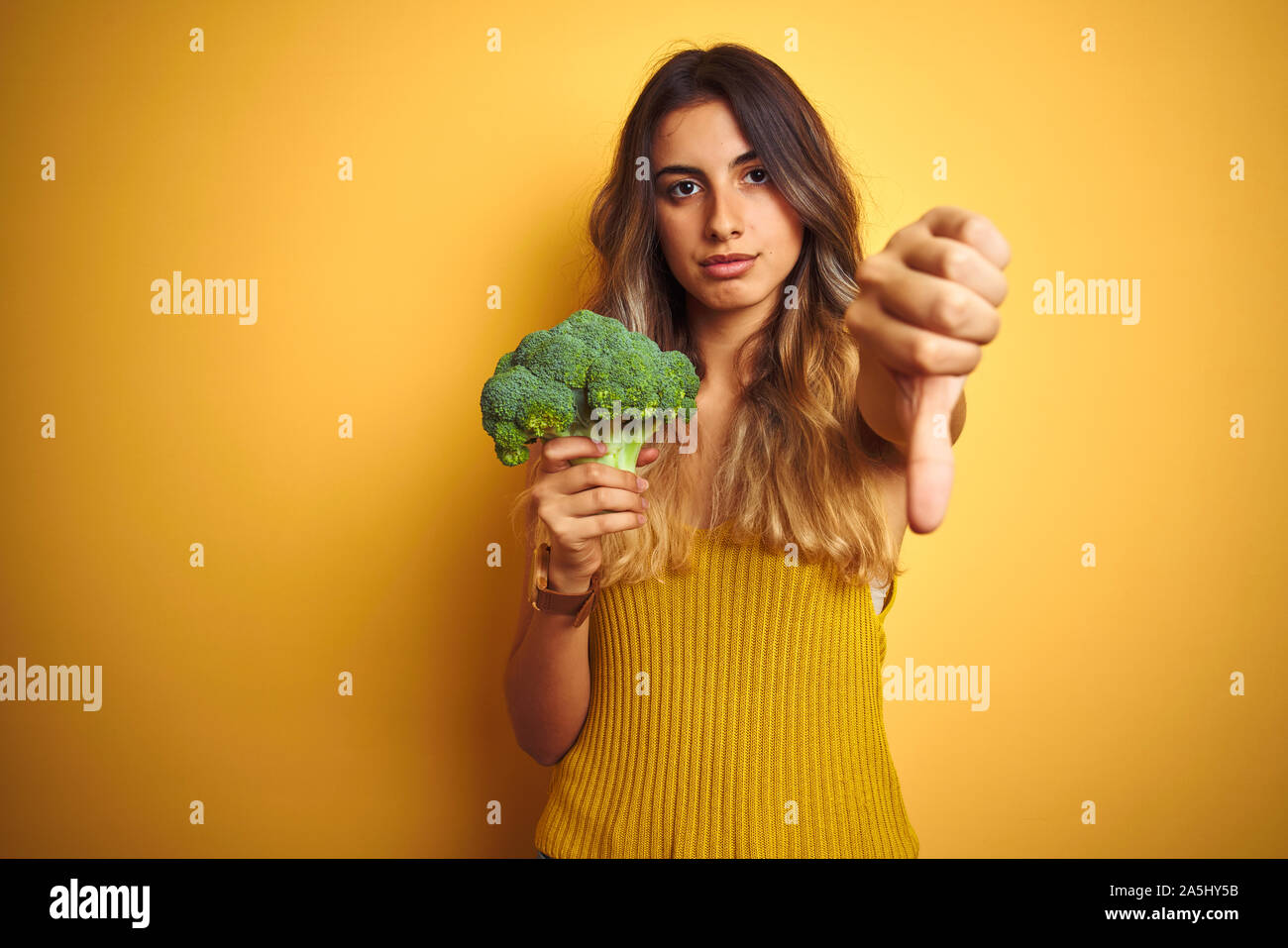 Young beautiful woman eating broccoli over yellow isolated background ...