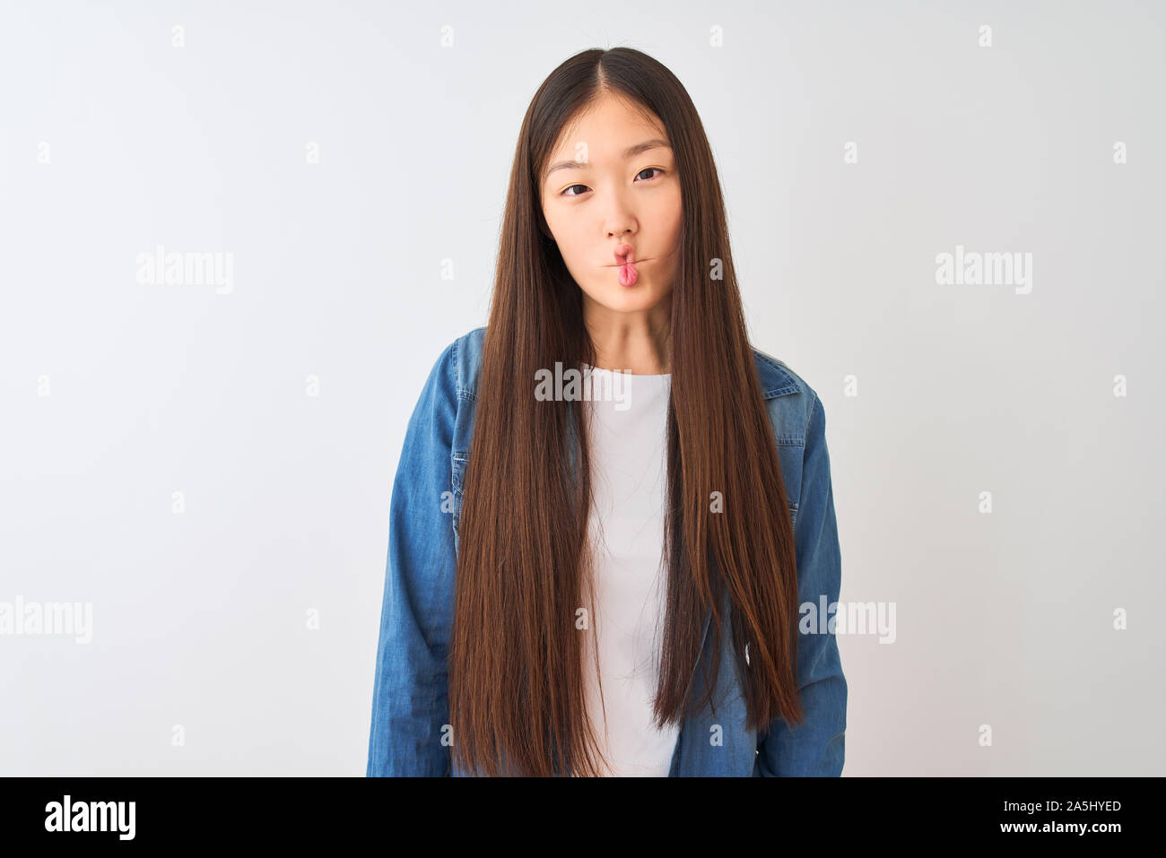 Young chinese woman wearing denim shirt standing over isolated white background making fish face ...