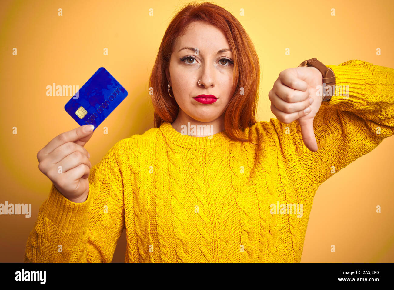 Young customer redhead woman holding credit card over yellow isolated ...