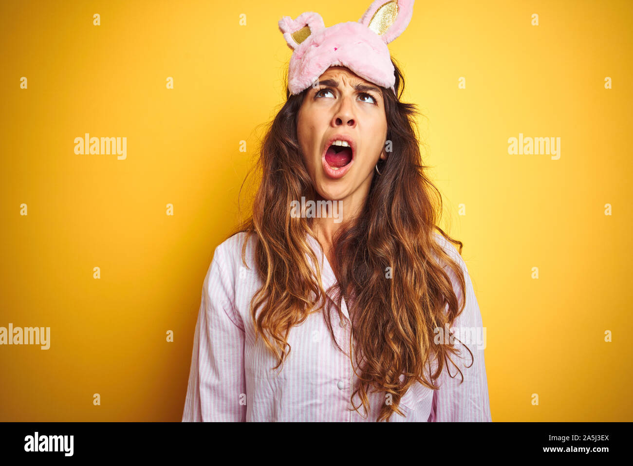 Young woman wearing pajama and sleep mask standing over yellow isolated ...