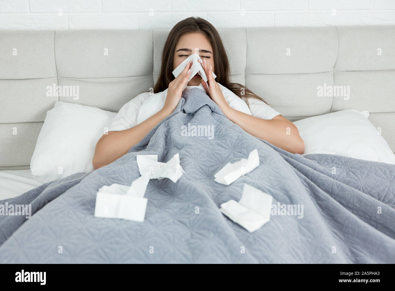young sick woman sitting in bed bowing out her running nose with paper ...