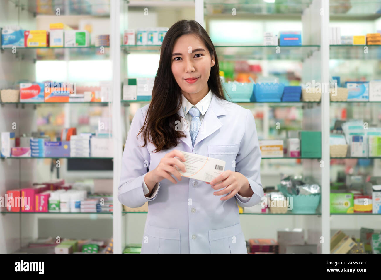 Asian young woman pharmacist with a lovely friendly smile holding medicinebox and looking at ...