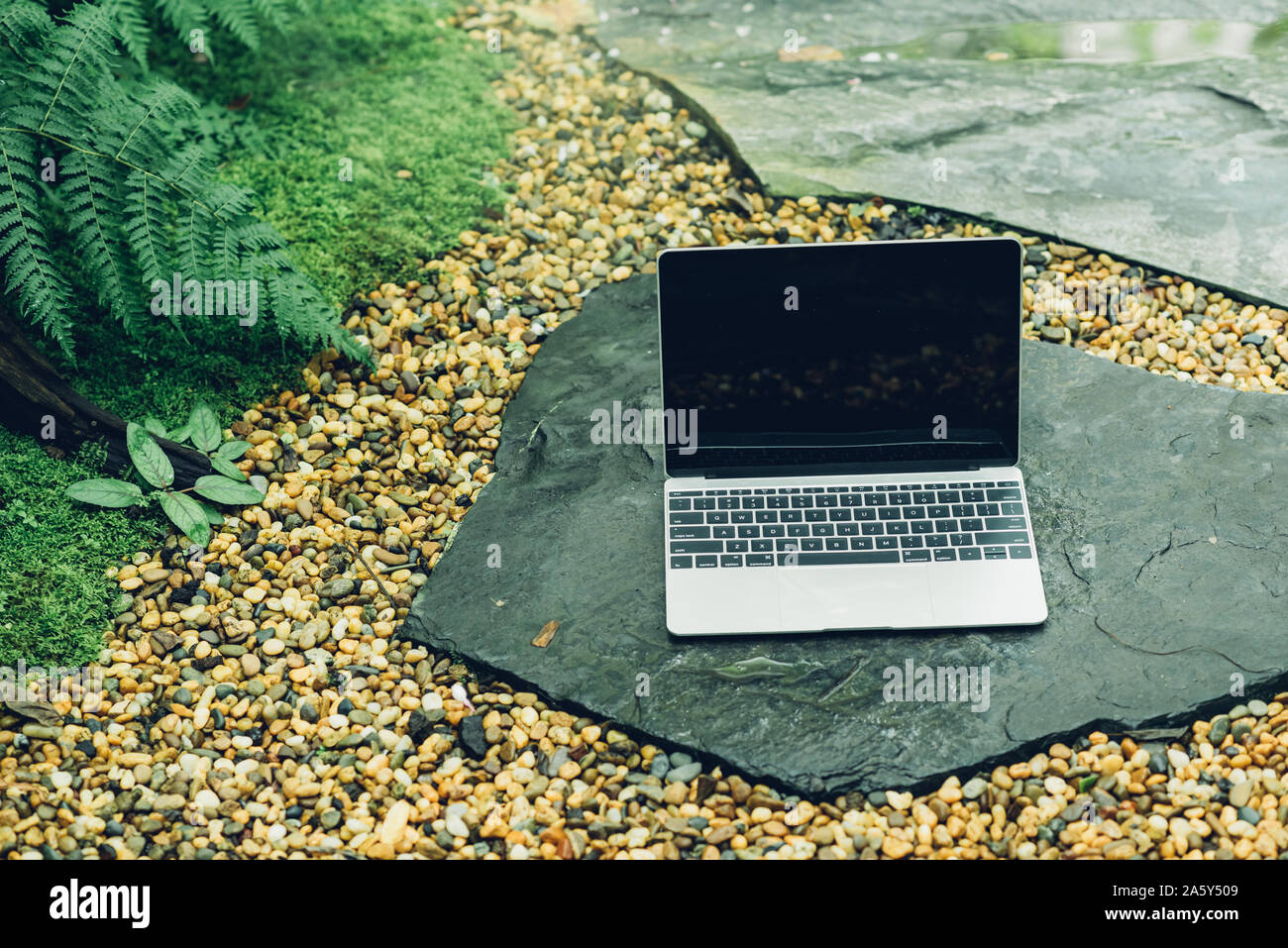 Laptop computer mockup has empty screen outside on stone in garden of ...