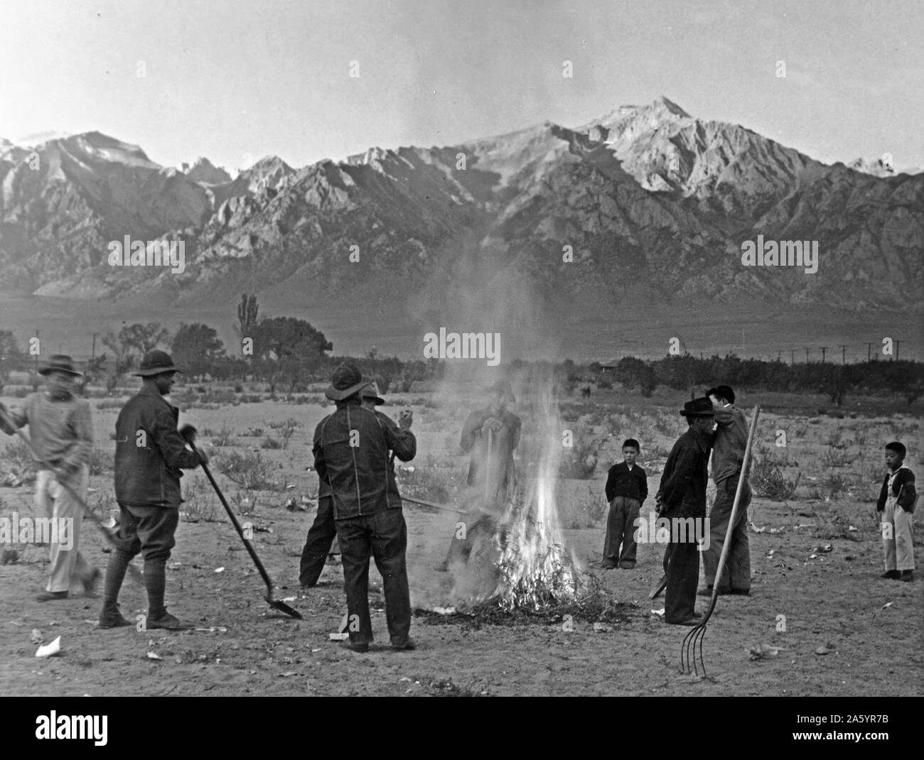 Photographic print of men and boys standing around a small brush fire ...