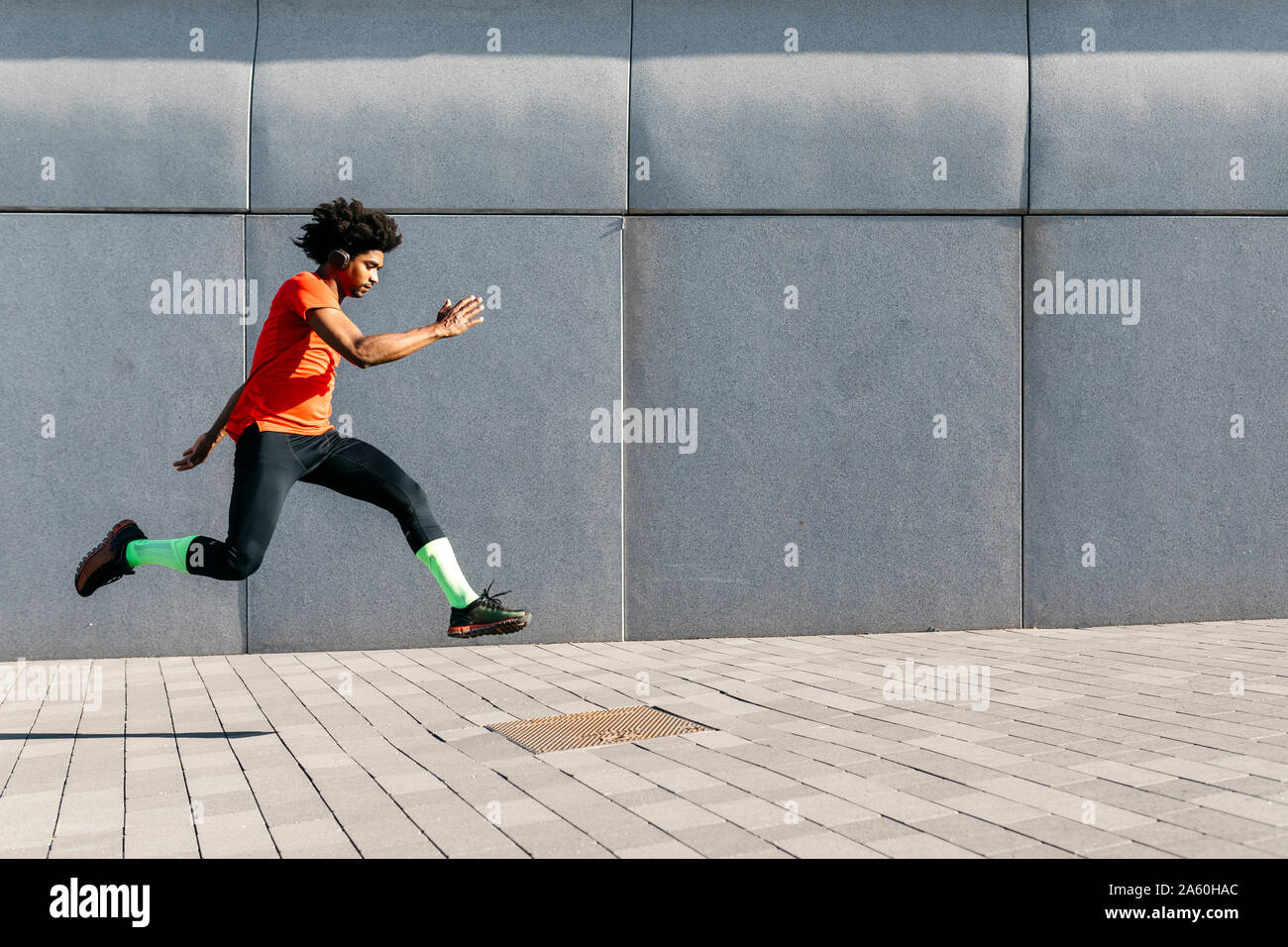 Young man jumping in front of a gray wall in the city, listening to ...