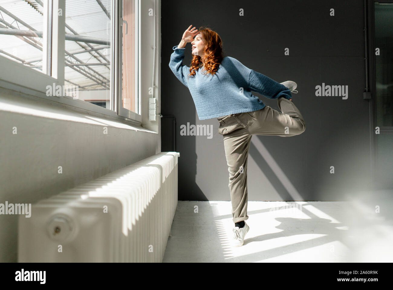 Redheaded woman standing on one leg in a loft looking out of window ...