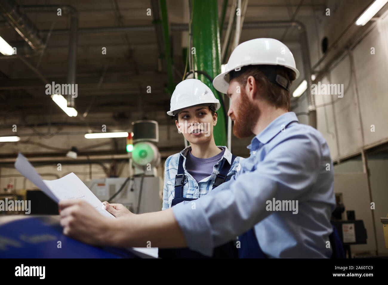 Young engineer in overalls and work helmets talking to her colleague ...