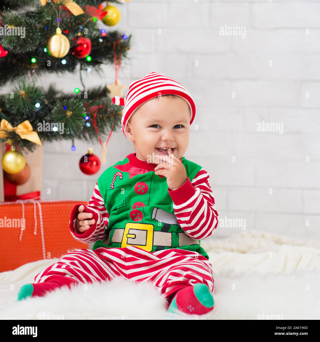 Cute baby elf enjoying sweet lollipop under Christmas tree Stock Photo ...