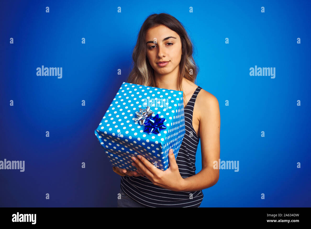 Young woman holding birthday present over blue isolated background ...