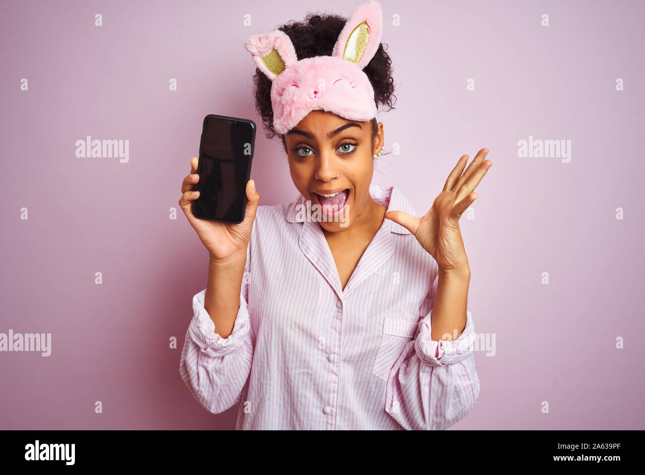 Afro woman wearing pajama and mask showing smartphone over isolated ...