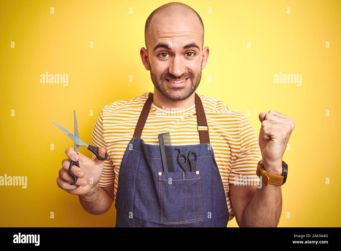Young hairdresser man wearing barber apron holding scissors over isolated yellow background ...