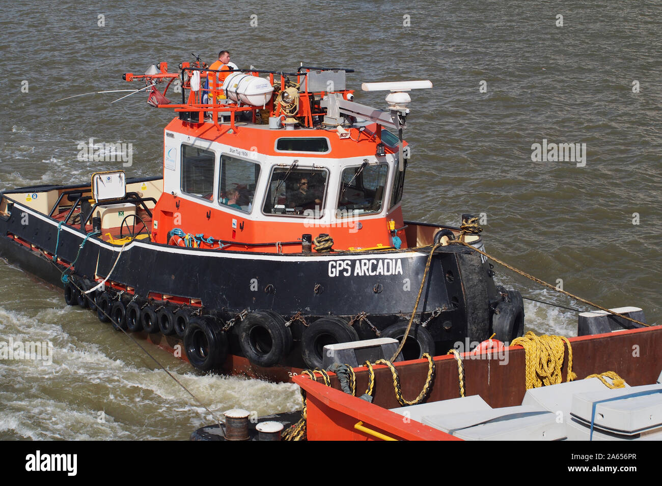 An orange and black tug boat on the River Thames pushing a working ...