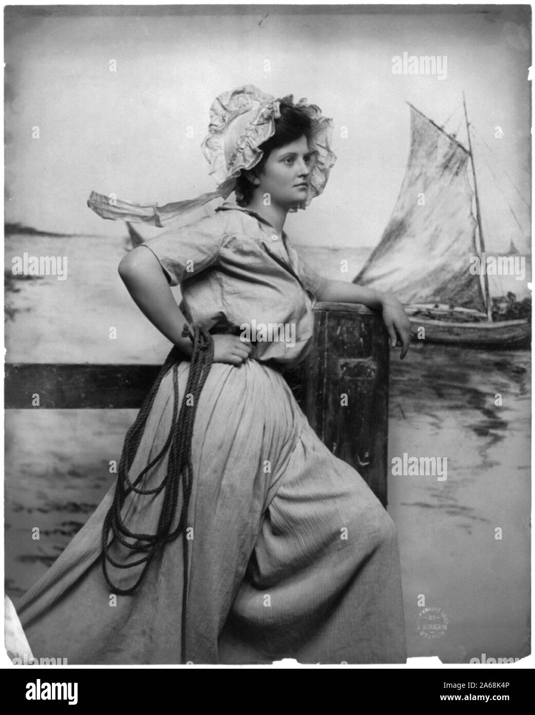 Woman wearing bonnet, holding rope and standing on pier; fishing boat ...