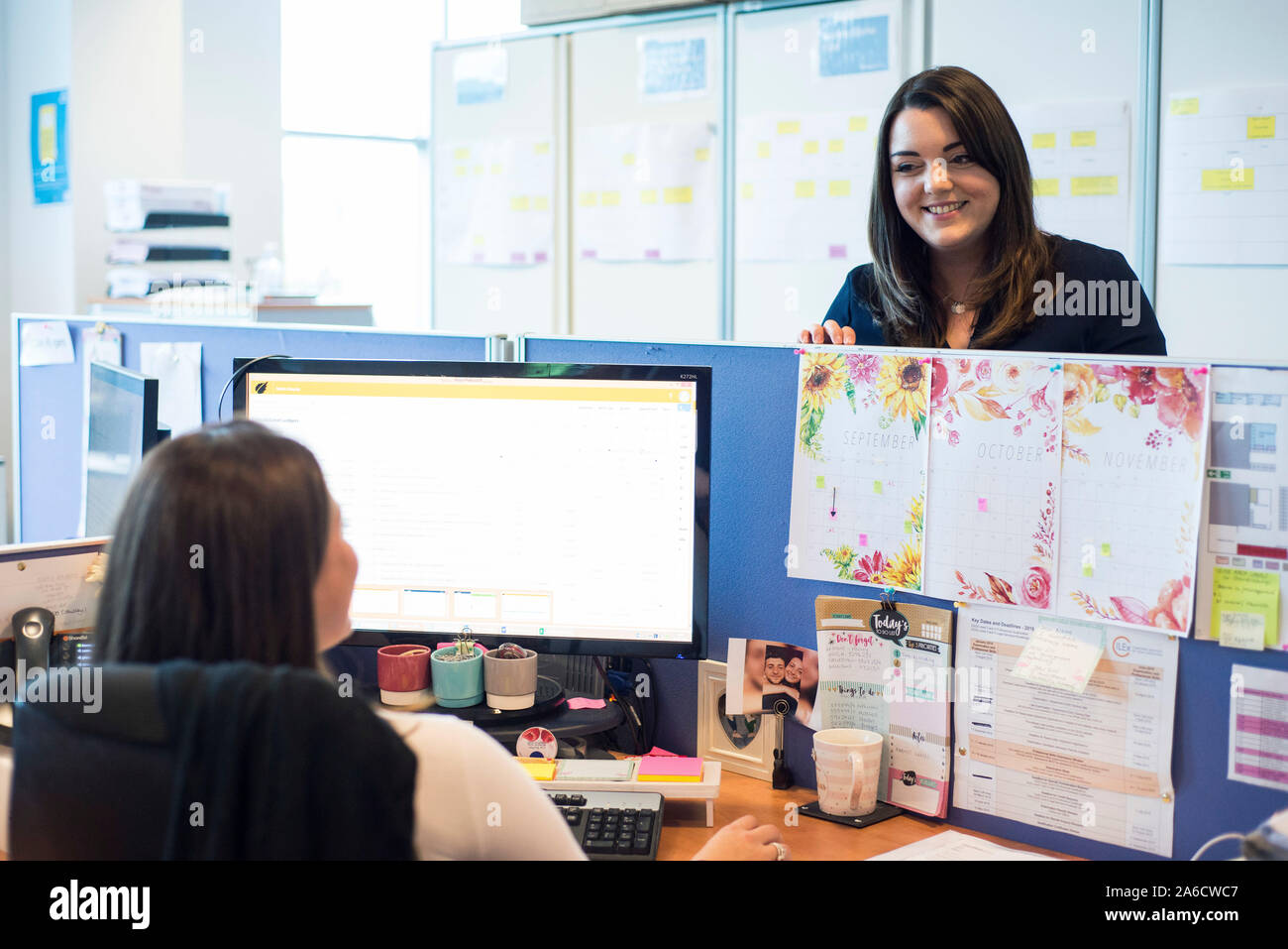 two female members of staff chat across an open plan office over the ...