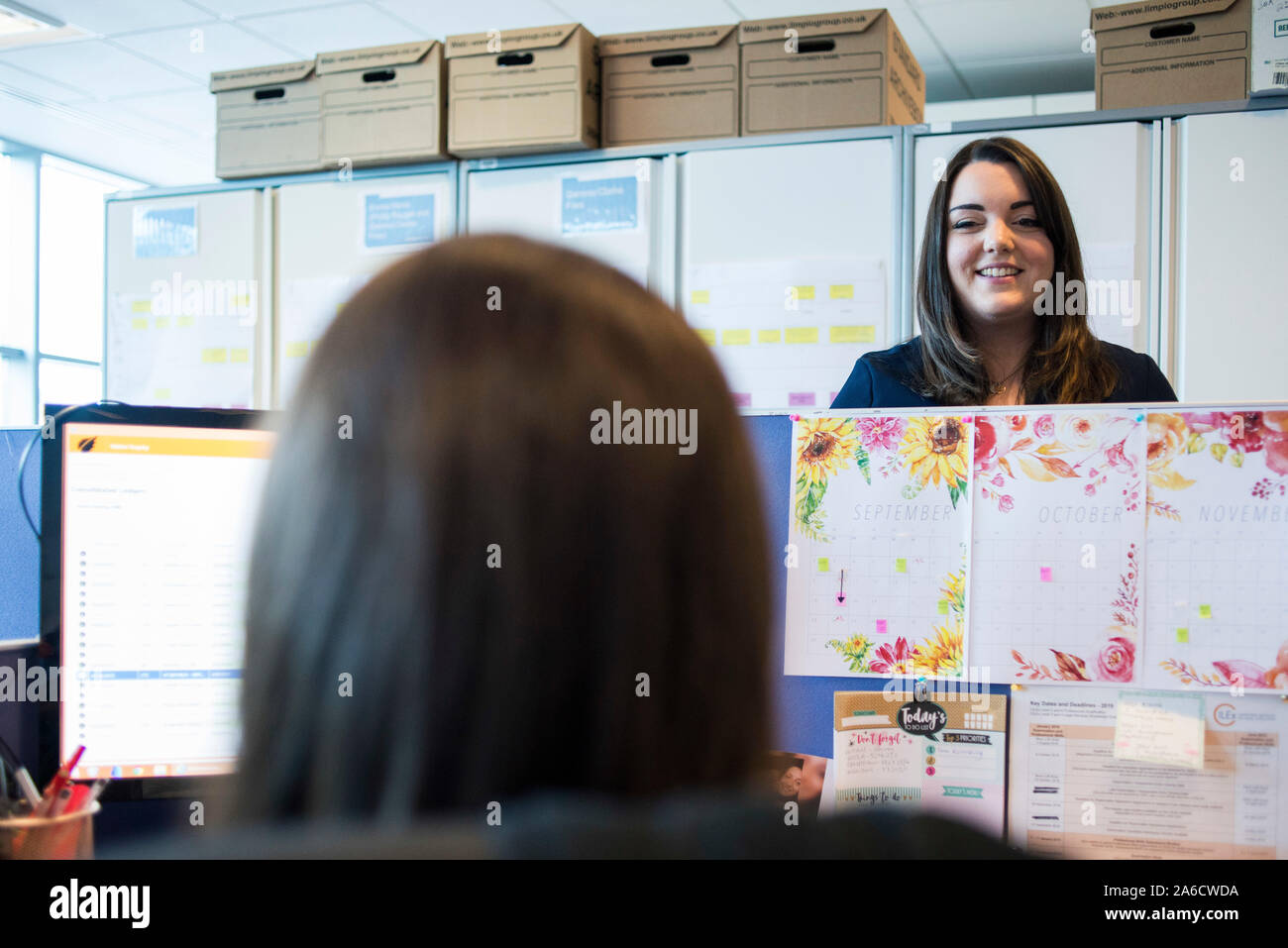 two female members of staff chat across an open plan office over the ...