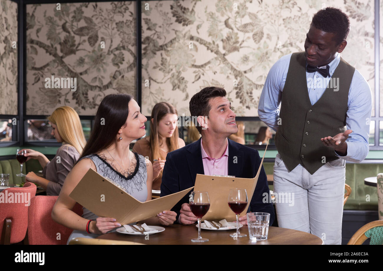 Polite African American waiter helping elegant couple with menu in ...