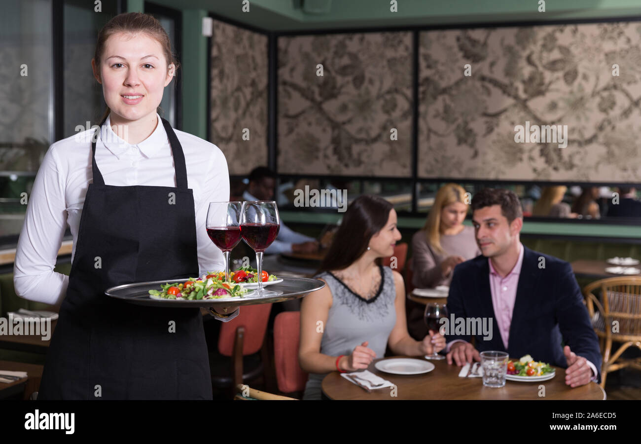 Polite waitress wearing apron holding tray with dishes in restaurant ...