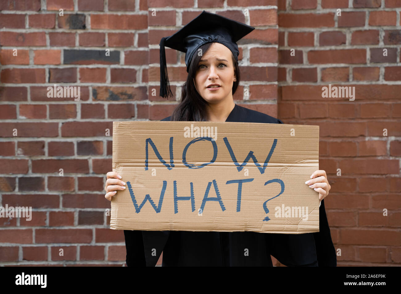 Sad Graduate Student Standing With Now What Placard Against Brick Wall ...