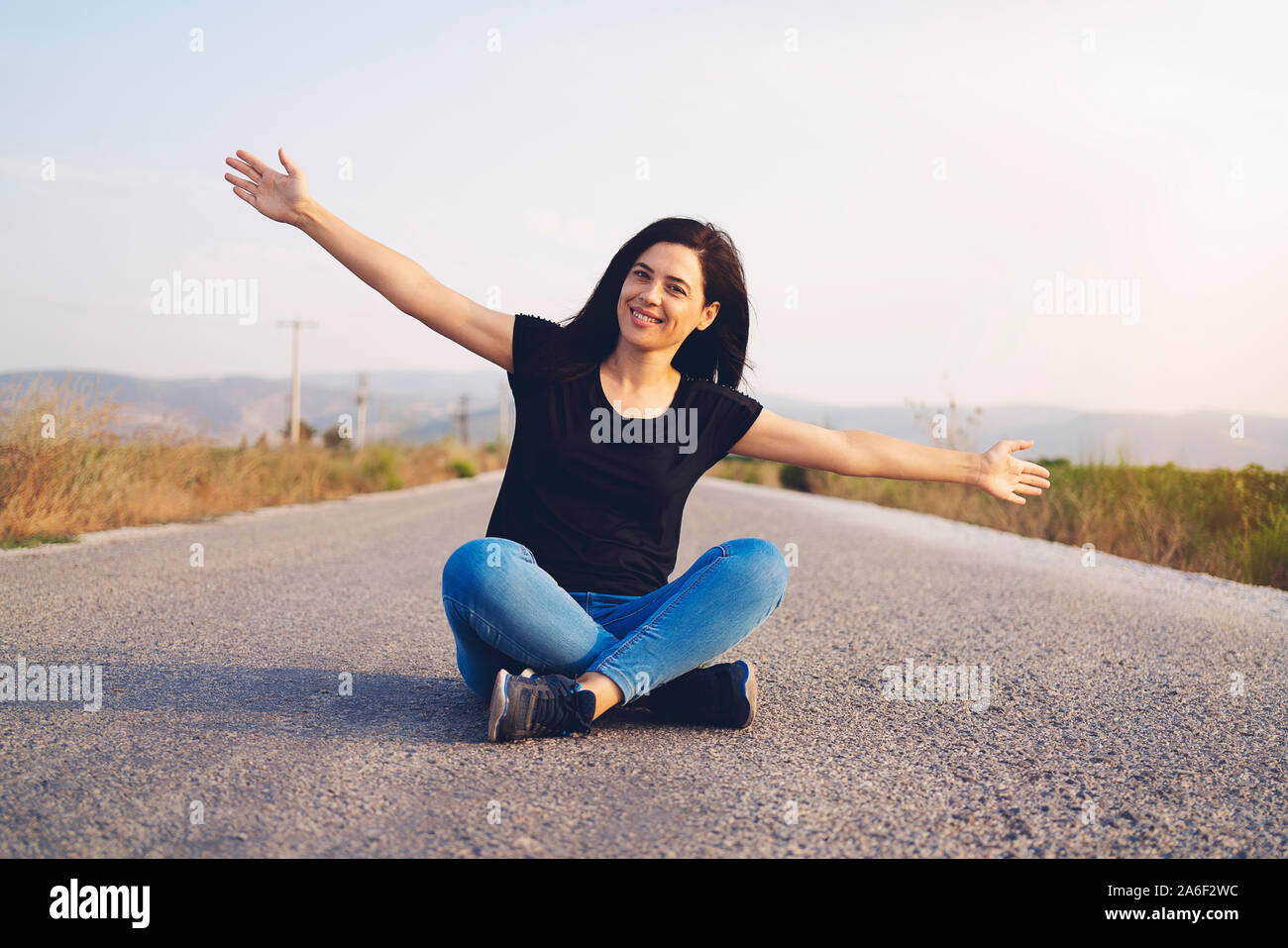 Happy woman standing with hands up on long straight road facing the sun ...